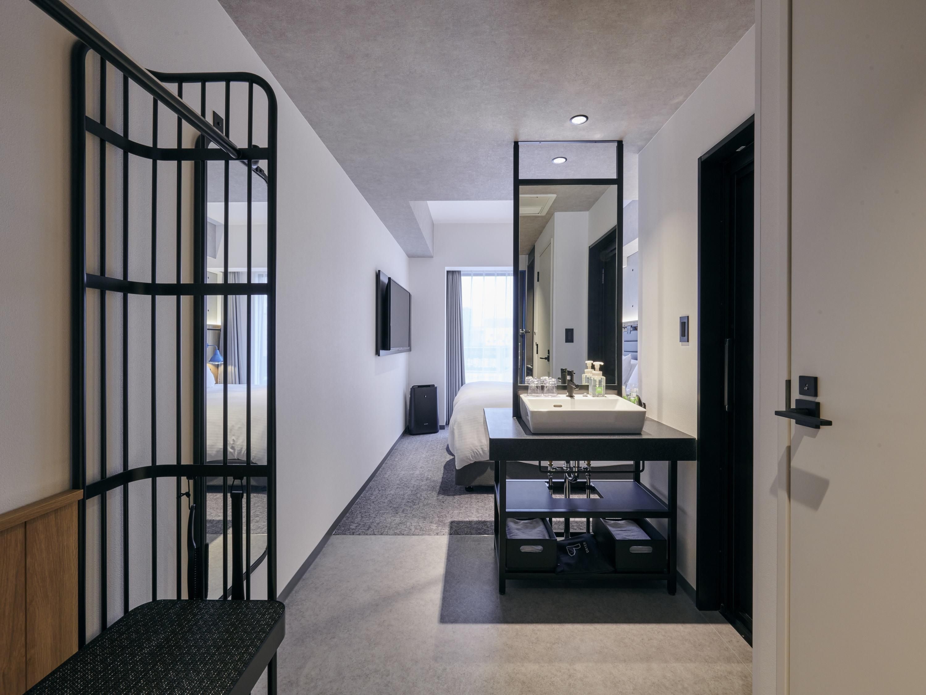 Quarto de hotel moderno com cama, escrivaninha, espelho e banheiro com pia e vaso sanitário.