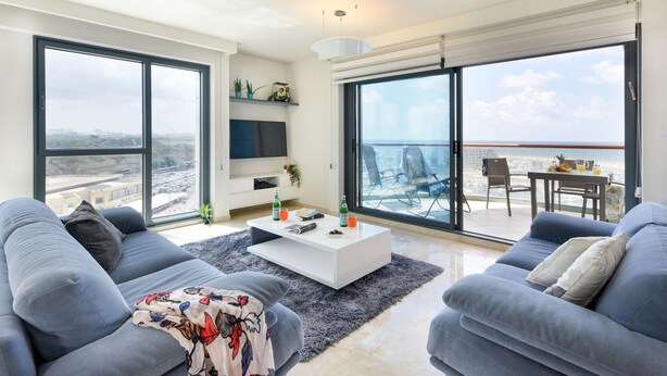 Sala moderna com um sofá grande, uma mesa de centro e uma TV. Há uma varanda com vista para a cidade.