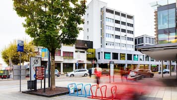 Um hotel moderno com fachada de vidro, um painel digital e área de estacionamento com barreiras azuis e vermelhas.