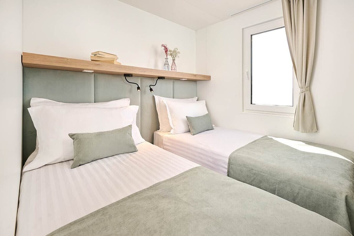 Quarto com duas camas, uma janela e uma estante com livros e flores.