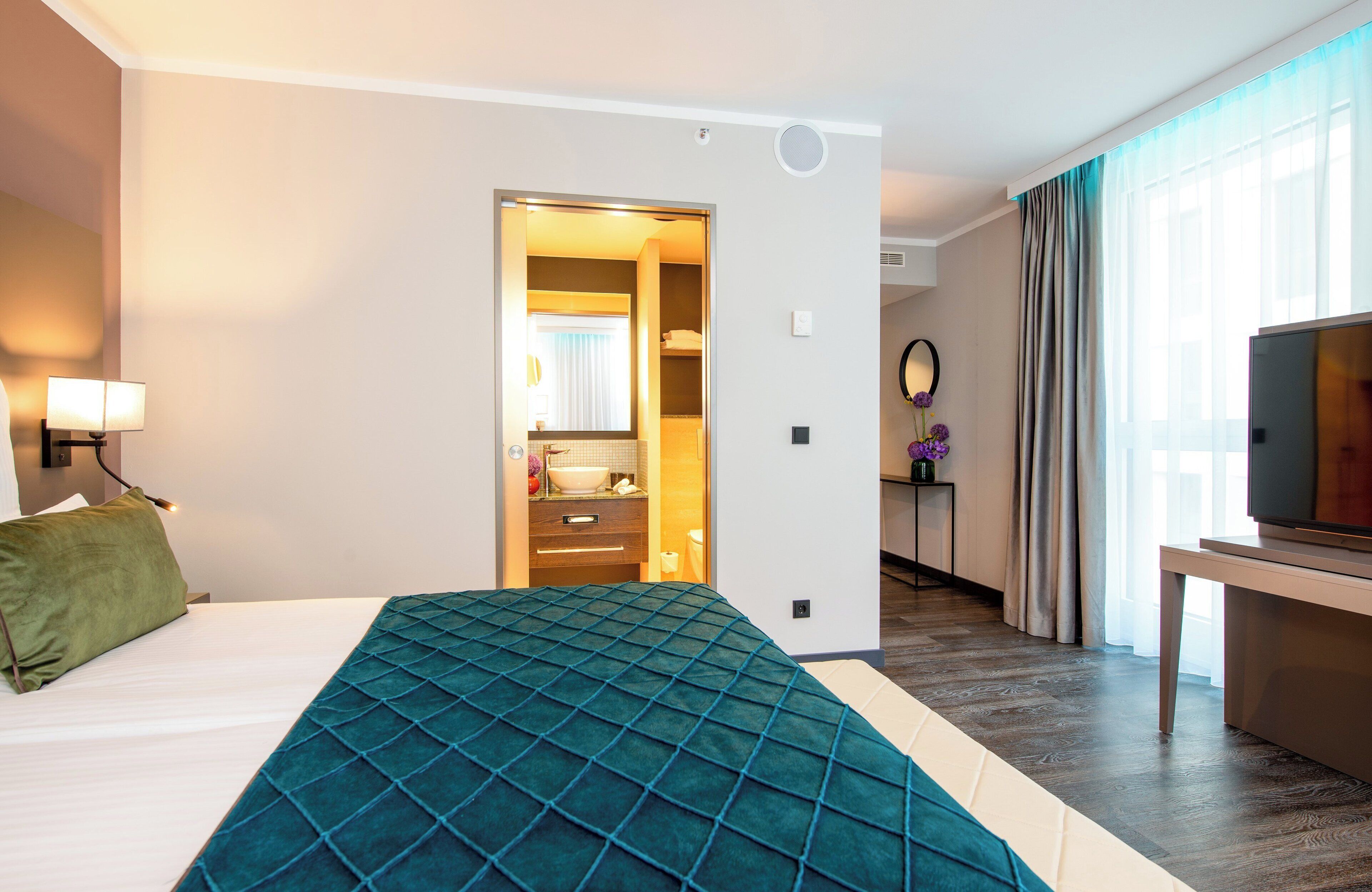 Quarto de hotel com cama, televisão, escrivaninha e banheiro visível através de uma porta aberta.