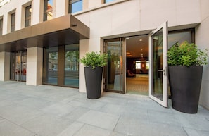 Entrada de um edifício moderno com portas de vidro, grandes vasos de plantas e um calçadão pavimentado.