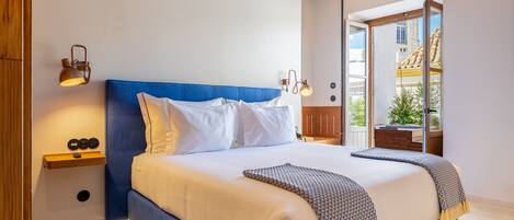 Cama arrumada com cabeceira azul, roupa de cama branca e manta decorativa estampada. Há duas mesas de cabeceira com abajures, uma pequena prateleira com objetos decorativos e vista para a varanda.
