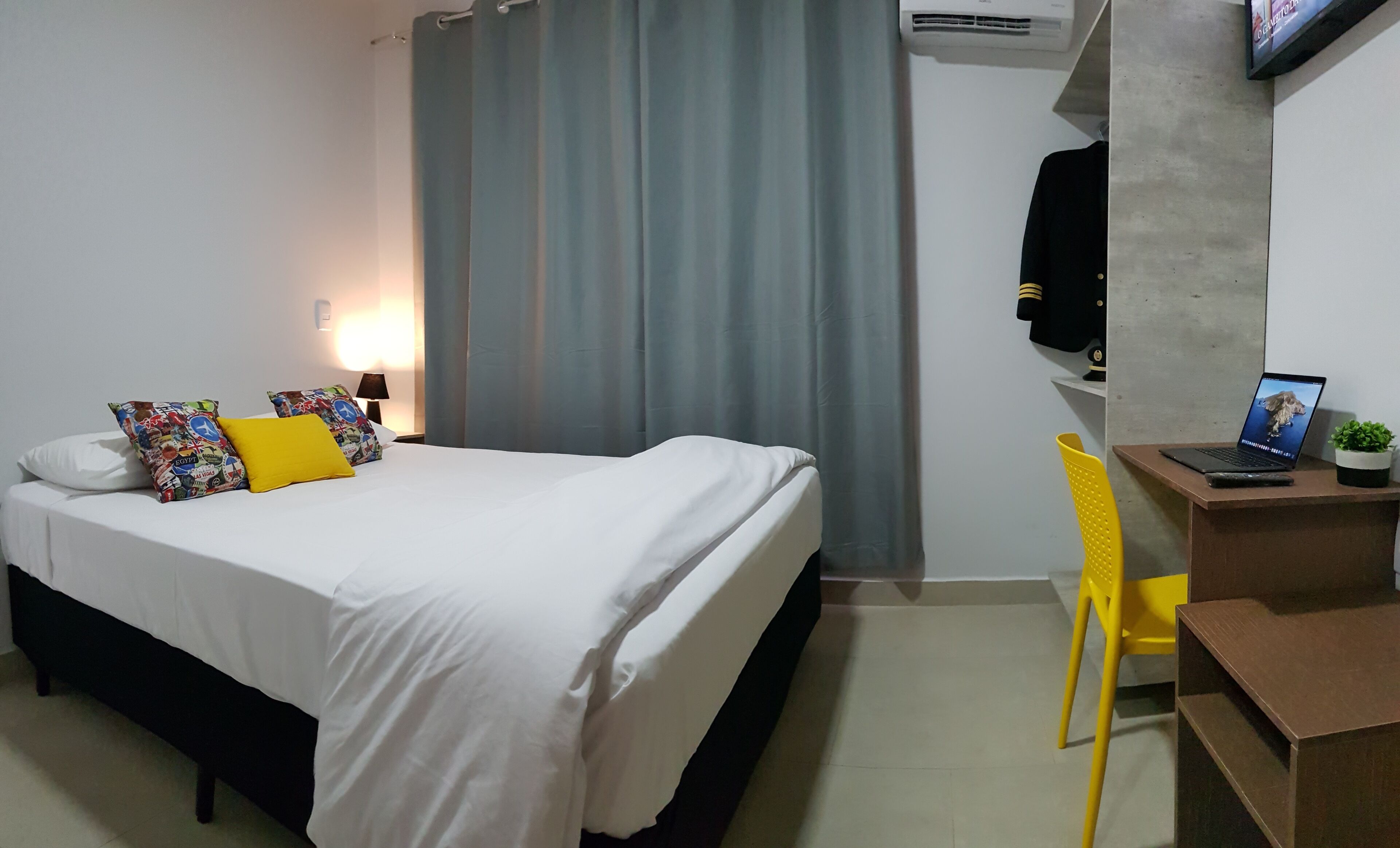 Cama bem arrumada com roupa de cama branca e almofadas coloridas, uma escrivaninha com um laptop e uma pequena planta, e uma cortina cinza ao fundo.