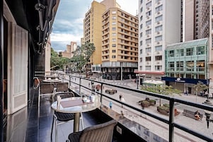 Uma área de estar ao ar livre com mesas e cadeiras, com vista para uma rua com edifícios e um café.