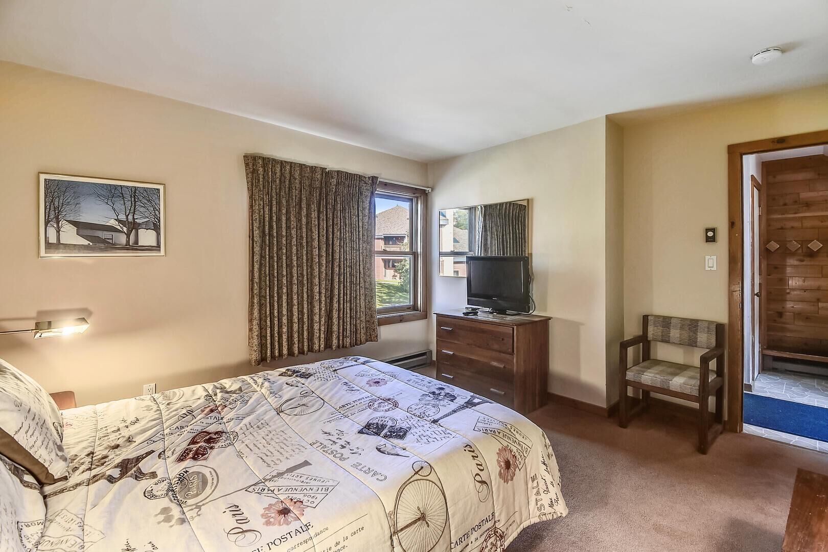 Quarto com uma cama, televisão sobre uma cômoda de madeira, uma cadeira e uma janela com cortinas.