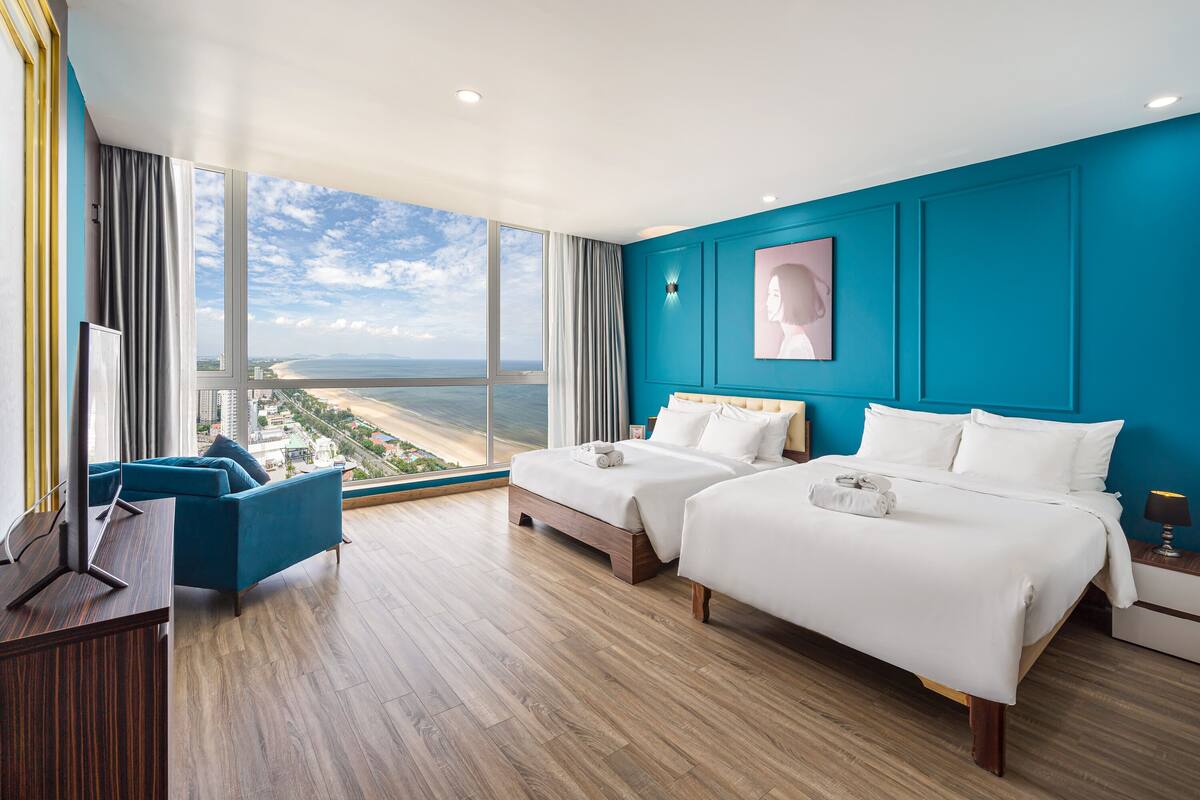 Quarto de hotel moderno com duas camas, uma janela grande com vista para a cidade, uma parede de destaque azul e piso de madeira.