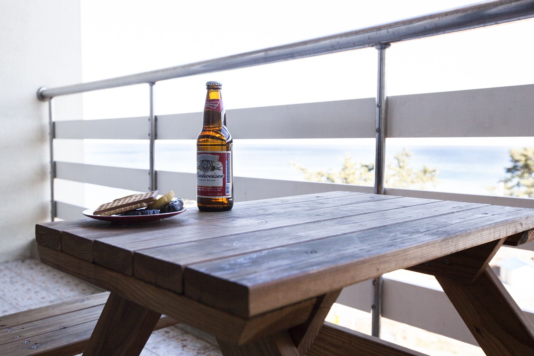 Uma mesa de madeira com uma garrafa de cerveja Budweiser e um prato de comida numa varanda com guarda-corpo.