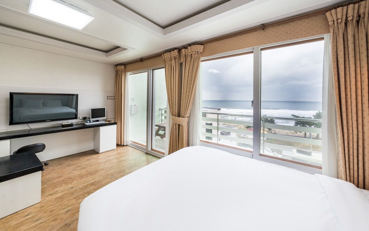 Quarto de hotel com cama, escrivaninha com computador, televisão e uma janela grande com cortinas.