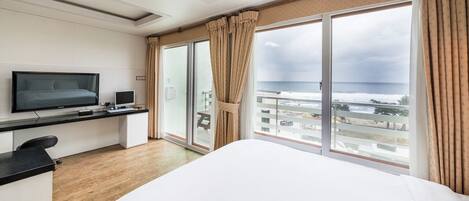 Quarto de hotel com cama, escrivaninha com computador, televisão e uma janela grande com cortinas.
