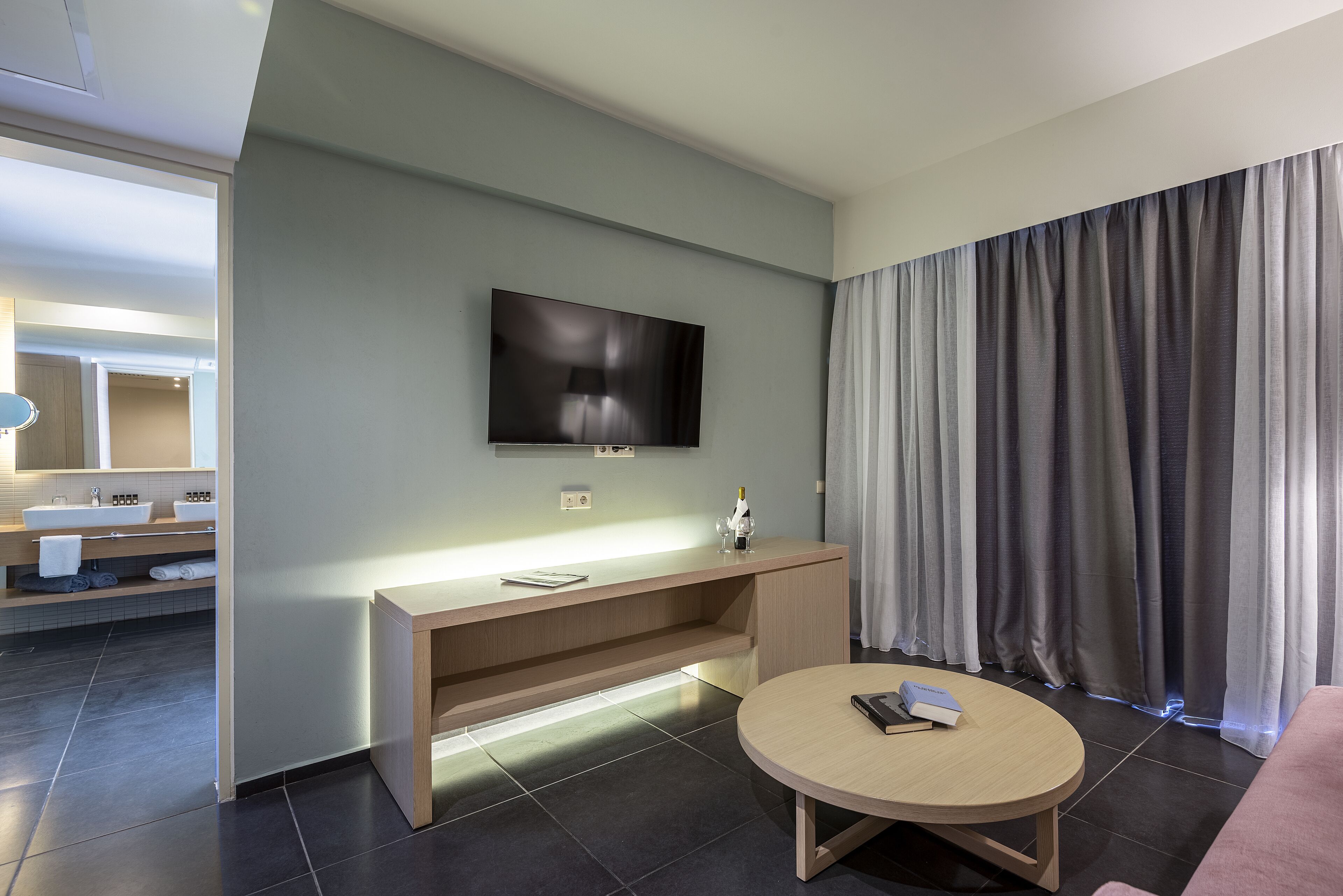 Quarto de hotel moderno com televisão de tela plana na parede, escrivaninha de madeira com luminária, mesa redonda e uma janela grande com cortinas.