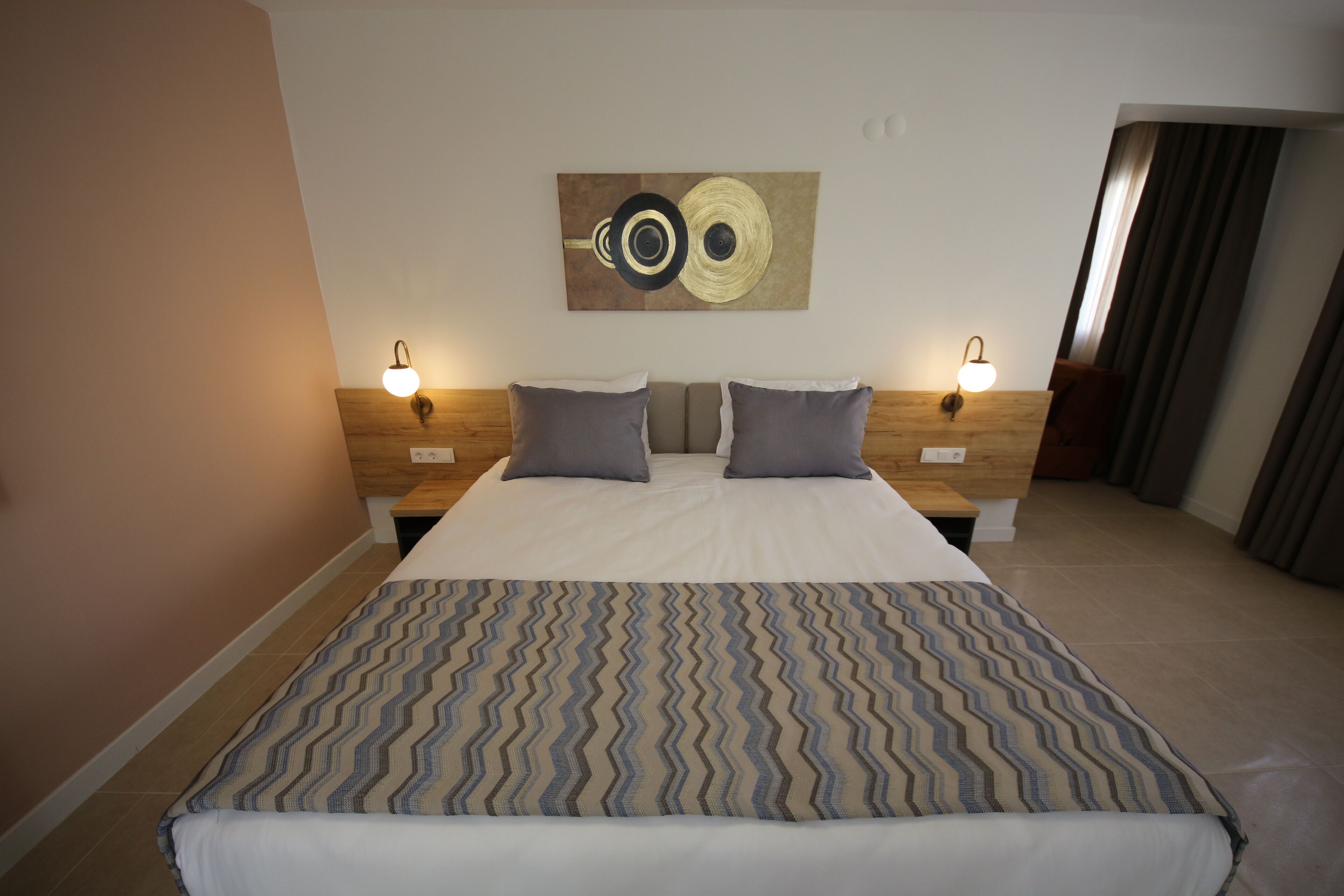 Quarto de hotel com uma cama grande, duas mesas de cabeceira, uma obra de arte na parede e uma janela com cortinas.