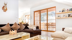 Sala de estar moderna com um sofá marrom, uma mesa de centro de vidro e um sofá bege. Há estantes de madeira com livros e quadros, e uma grande janela com vista para móveis de exterior.