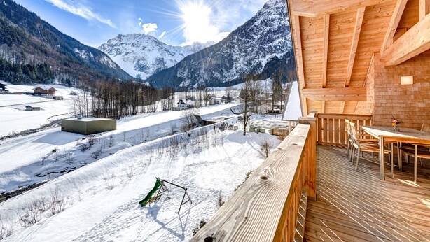 Um deck de madeira com mesa e cadeiras, com vista para uma paisagem montanhosa coberta de neve e céu claro.