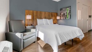 Quarto de hotel com cama, poltrona, mesa de cabeceira, luminária e pintura de uma paisagem urbana.