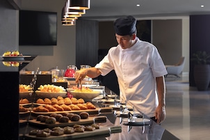 Um chef com uniforme branco e chapéu preto prepara comida em uma cozinha moderna com sistema de buffet.