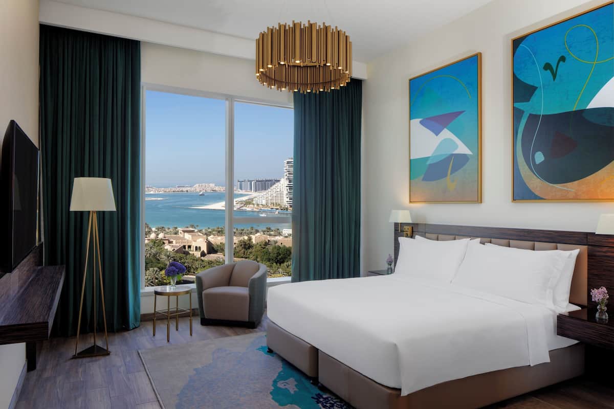 Quarto de hotel com uma cama grande, uma cadeira, uma escrivaninha, uma televisão e vista para o oceano e edifícios.
