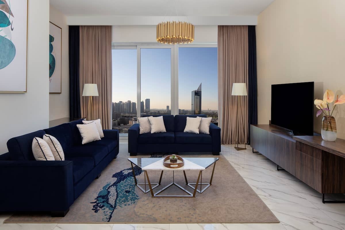 Uma sala de estar moderna com dois sofás azul escuro, uma mesa de centro de vidro, uma TV de tela plana e vista para a cidade através de janelas amplas.