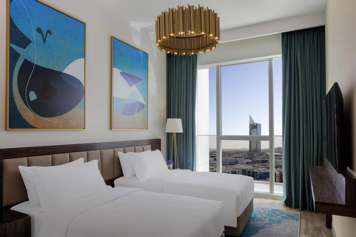 Quarto de hotel com duas camas, uma janela grande com vista para a cidade, quadros abstratos na parede e um lustre moderno.
