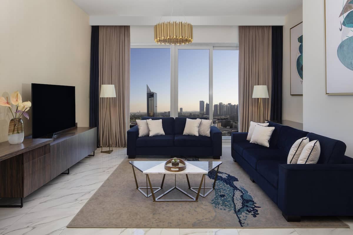 Uma sala de estar moderna com uma televisão grande, sofás azul escuro, uma mesa de centro de vidro e vista para a cidade através de amplas janelas.