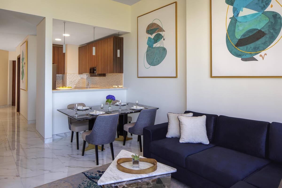 Uma sala de estar moderna com um sofá azul escuro, um espaço de jantar com mesa de vidro e uma cozinha ao fundo.