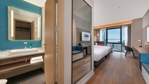 Quarto de hotel com banheiro, cama, escrivaninha e uma grande janela com vista para a cidade.