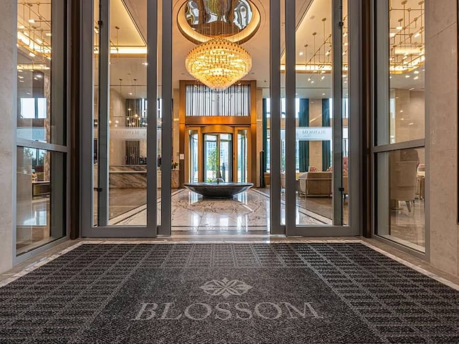Um saguão de hotel com um lustre imponente, uma recepção e um tapete com o nome do hotel.