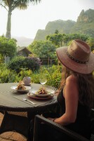 Uma pessoa sentada em uma mesa ao ar livre, com pratos de comida e uma taça de vinho, com vista para um jardim exuberante e paisagem montanhosa.
