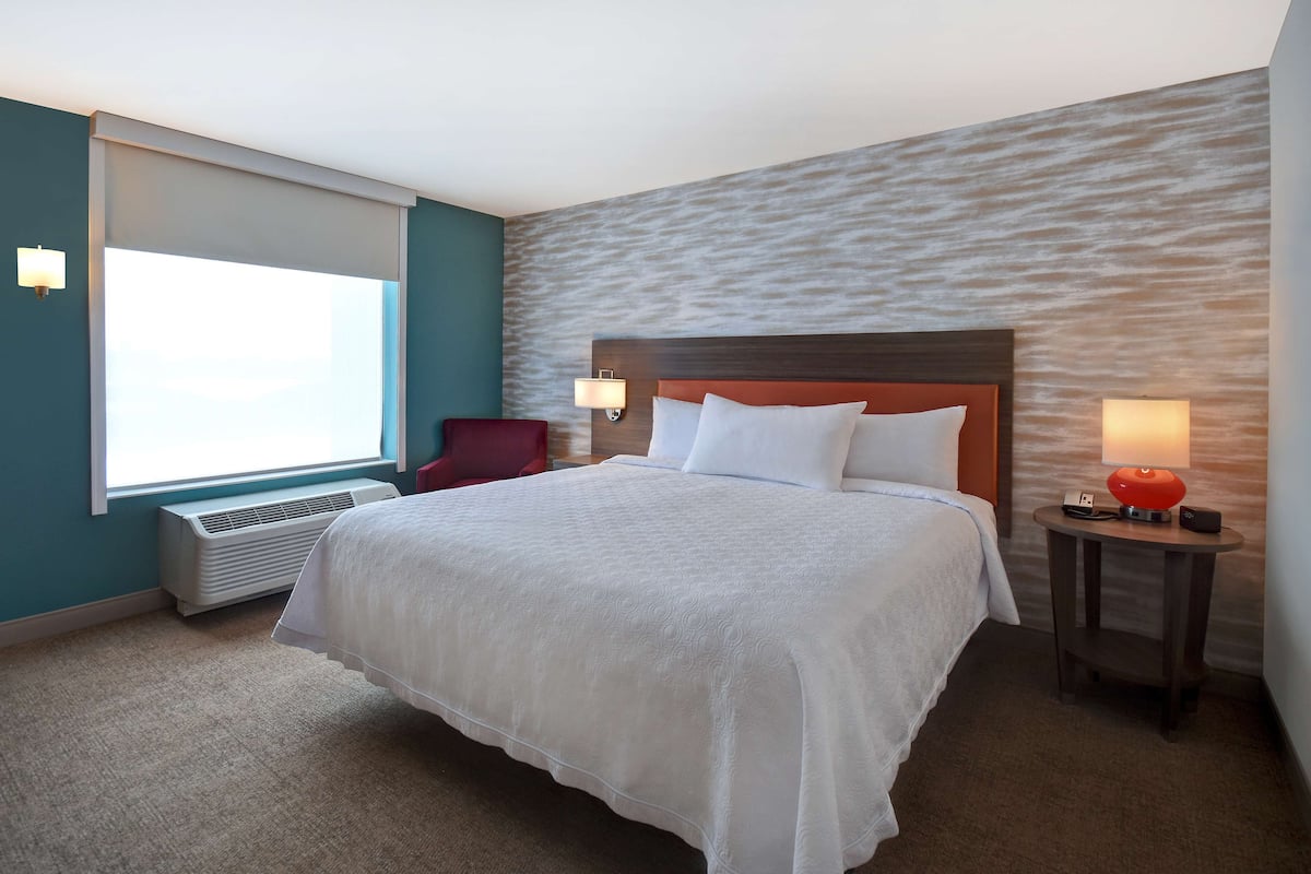 Quarto de hotel com uma cama grande, duas luminárias de cabeceira, uma cadeira vermelha e uma janela com persianas.