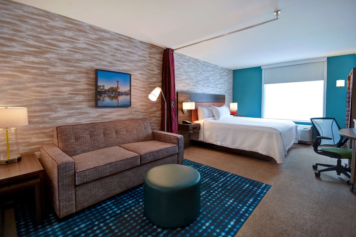 Quarto de hotel com sofá, um banquinho redondo, cama, escrivaninha com cadeira e janela com cortinas.