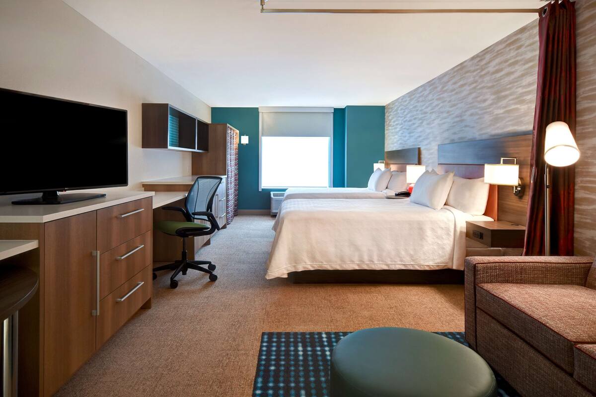 Quarto de hotel com uma cama, uma secretária com televisão de ecrã plano, uma cadeira, um candeeiro de mesa e uma janela com cortinas.