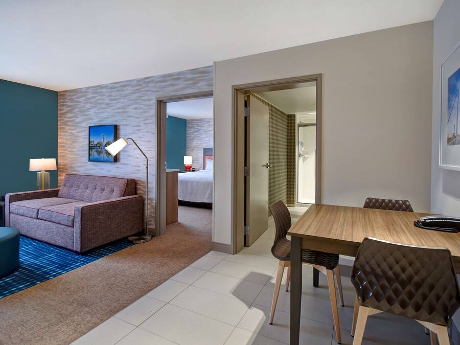 Quarto de hotel com sofá, mesa de jantar com cadeiras, luminária e cama ao fundo.