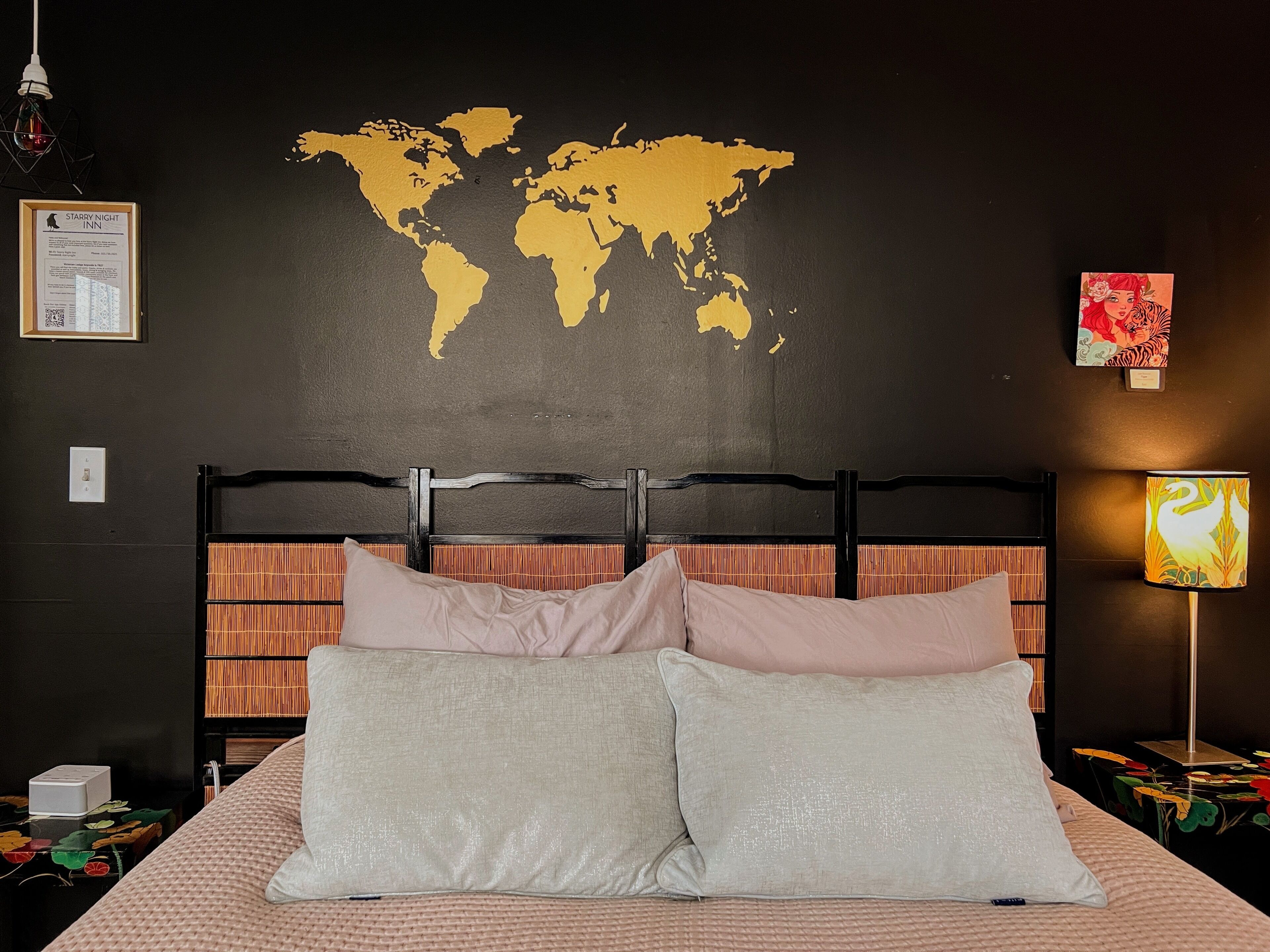 Quarto de hotel com cama, almofadas, um mapa-múndi na parede, um certificado emoldurado e uma luminária.