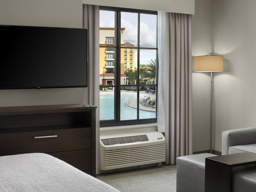 Quarto de hotel com cama, televisão, janela com vista para uma piscina e edifícios, luminária e cadeira.