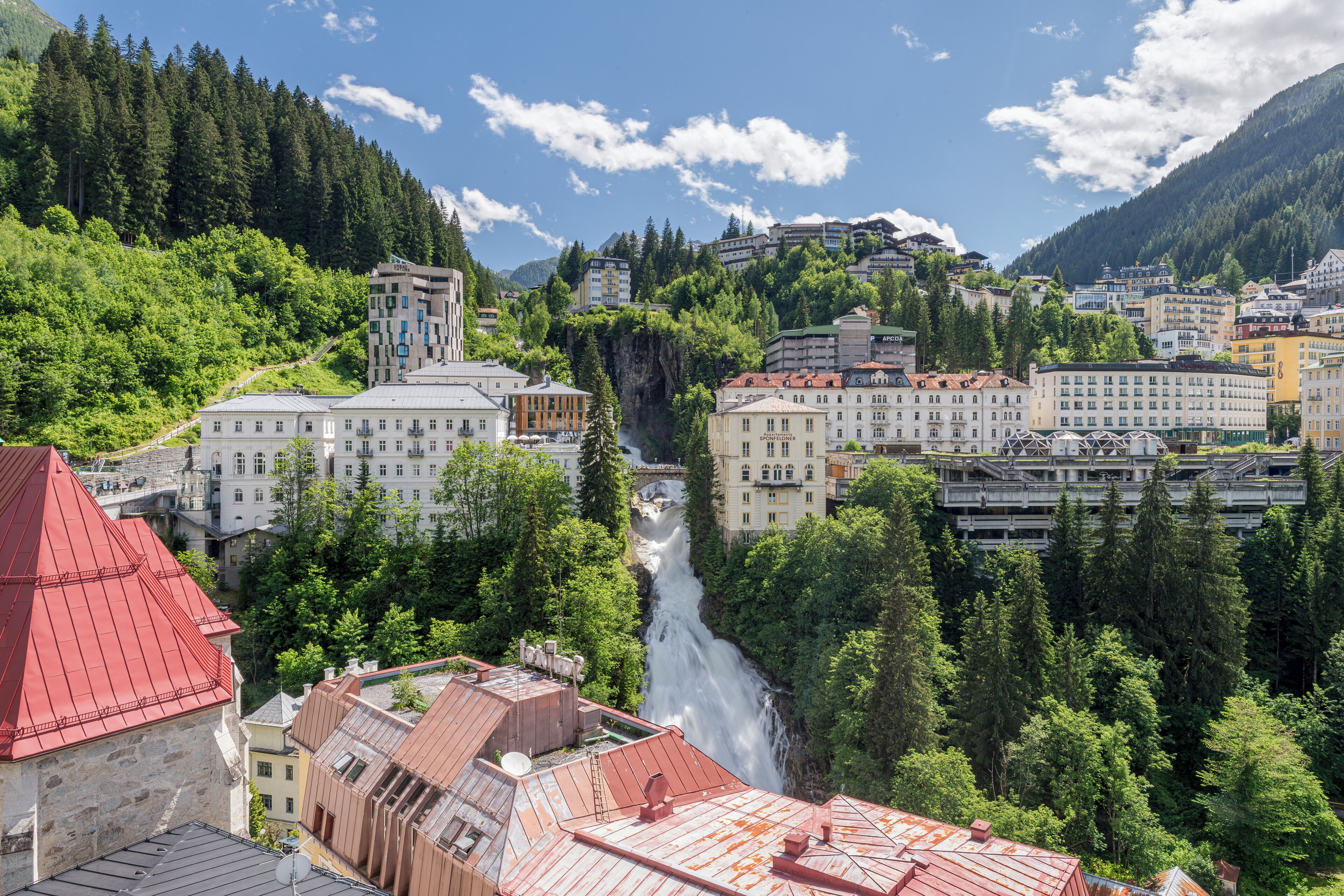Uma cidade com edifícios, uma cascata e colinas arborizadas.