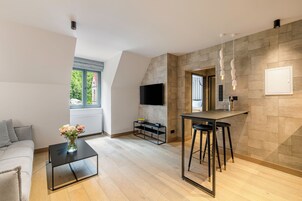 Uma sala de estar moderna com um sofá, uma mesa de centro com um vaso, uma TV fixada numa parede de pedra, e uma área de cozinha com um banco alto.
