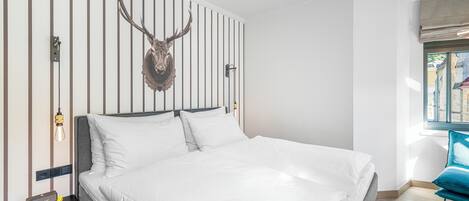 Quarto com cama, cabeça de veado montada na parede, mesa de cabeceira com livros e janela com vista.