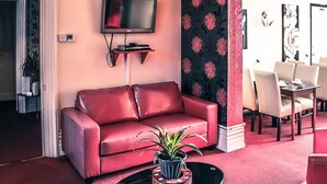 Uma sala de estar com um sofá de couro vermelho, uma mesa de centro preta com uma planta, uma TV de tela plana fixada na parede e um espaço de jantar com cadeiras e uma mesa.
