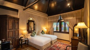 Quarto de hotel com duas camas, um guarda-roupa de madeira, uma mesinha com abajur, uma cadeira e uma janela grande com cortinas.