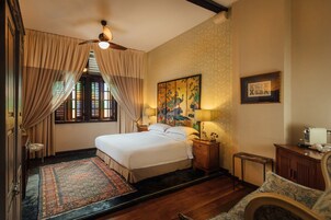 Quarto de hotel com uma cama grande, piso de madeira, tapete tradicional, escrivaninha com luminária e um quadro na parede.