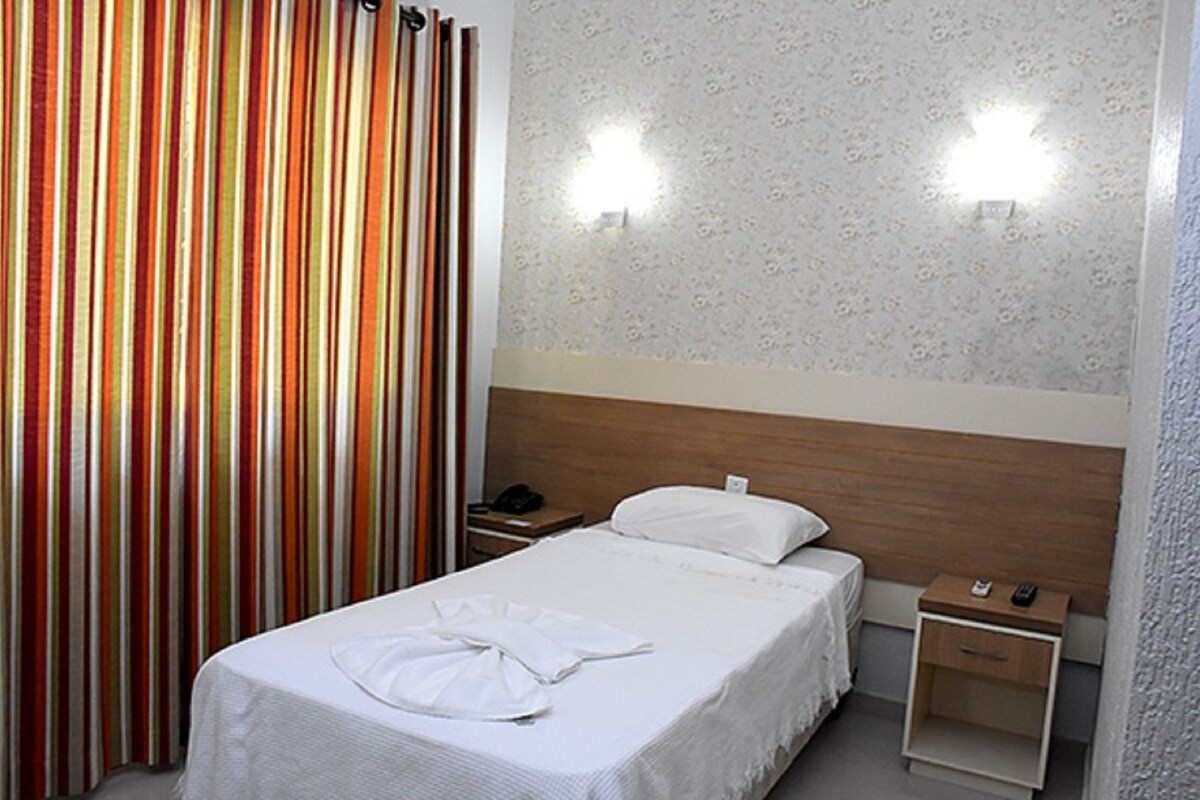 Quarto de hotel com cama, mesa de cabeceira, telefone e luminária na parede.