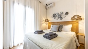 Cama bem arrumada com edredom branco e duas toalhas dobradas, cabeceira de madeira, ar condicionado fixado na parede e arte decorativa na parede.
