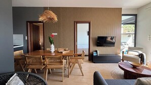 Uma sala de estar moderna com uma mesa de jantar de madeira, cadeiras de vime, uma TV de tela plana na parede e vista para o exterior através de uma grande janela.