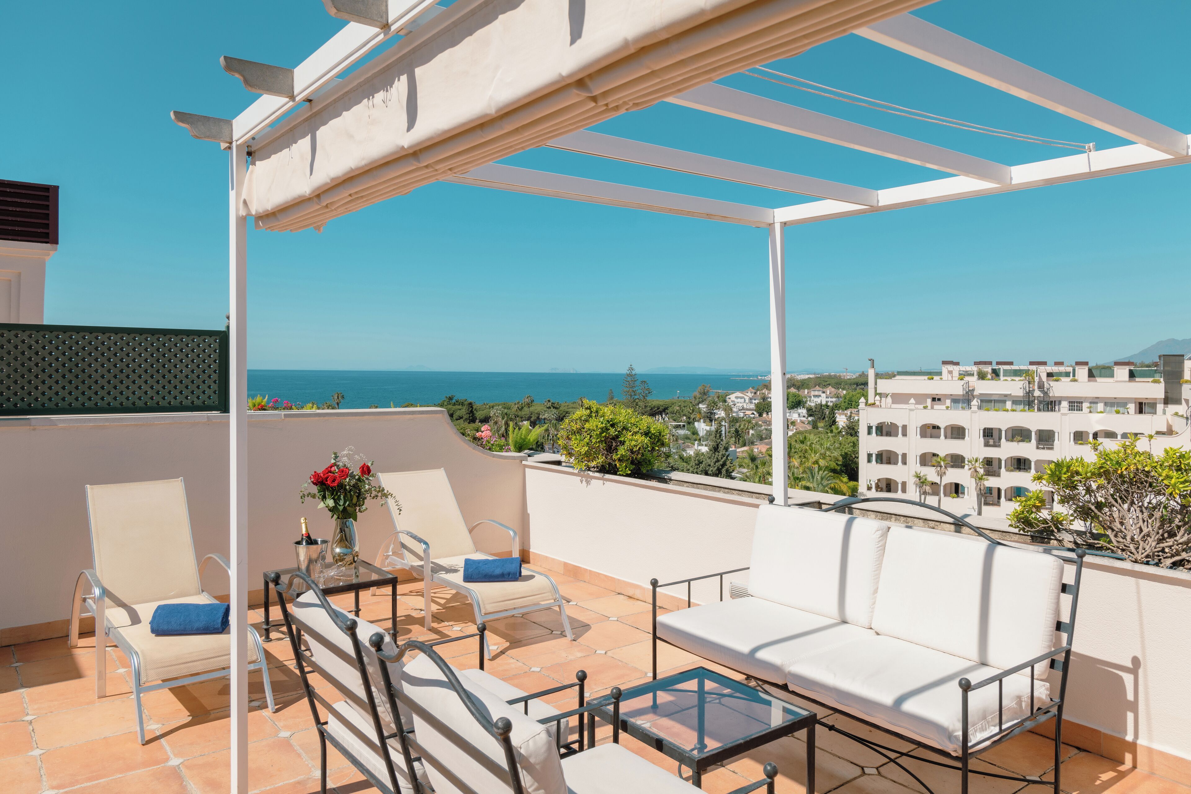 Terraço com sofá branco, duas poltronas e mesa de vidro, com vista para a cidade e o mar.