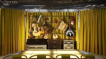 Quarto com cortina amarela ao fundo, parede com instrumentos musicais e uma mesa com copos e um alto-falante.