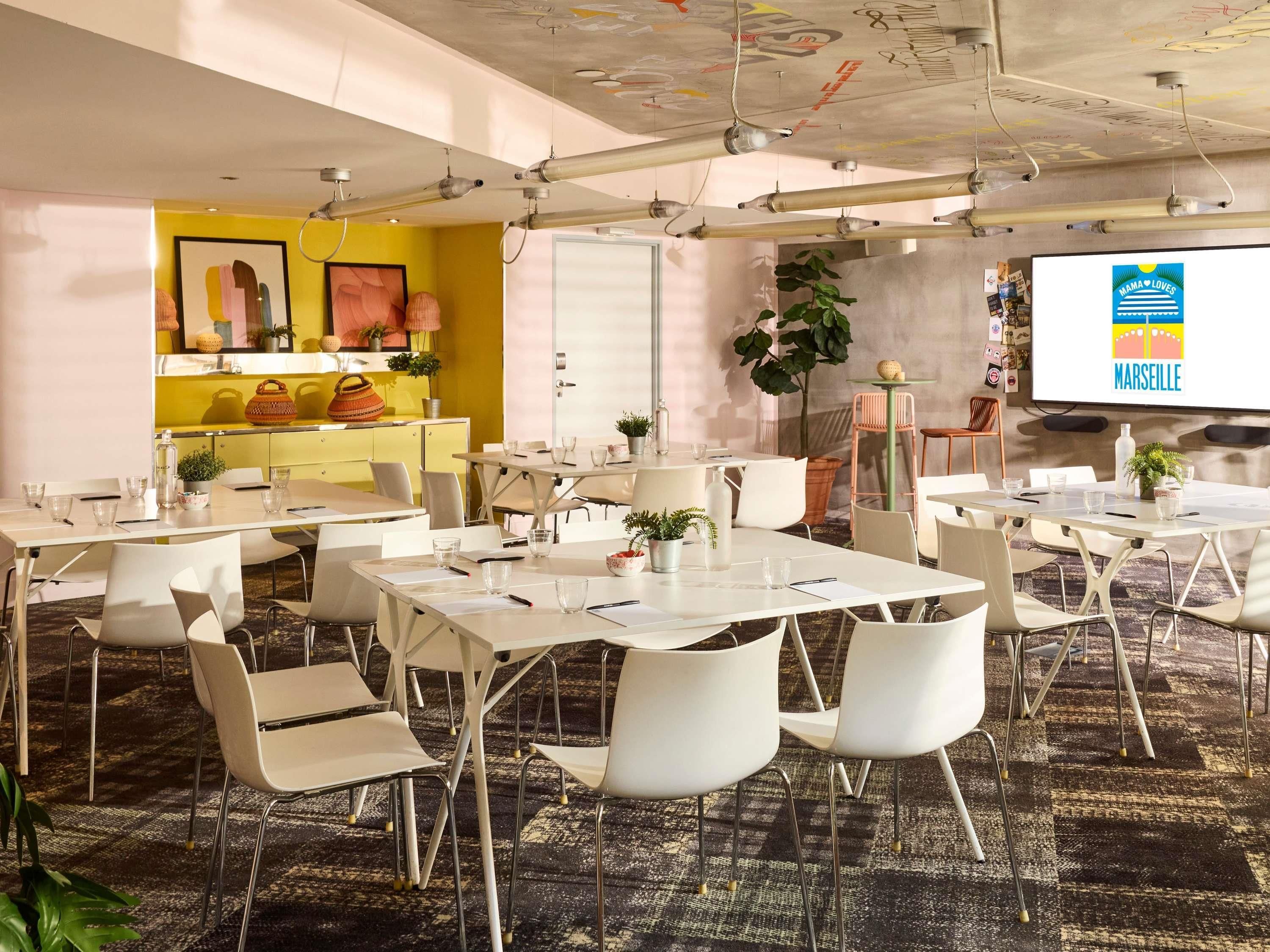 Uma sala de conferências com várias mesas e cadeiras, uma tela de projetor exibindo 'NAMA LOVES MARSEILLE' e uma parede amarela com objetos decorativos.