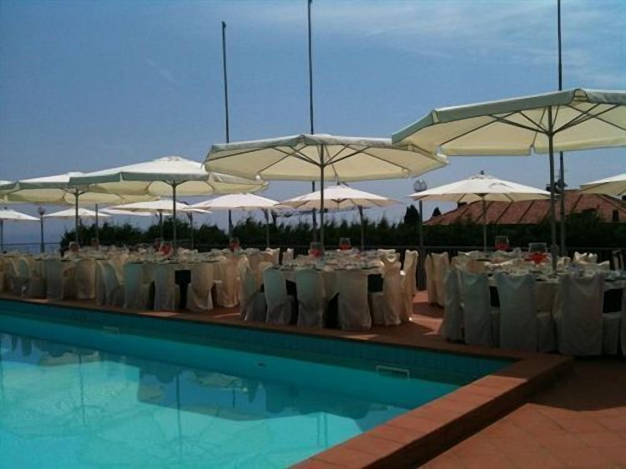 Uma área à beira da piscina com mesas preparadas para refeições, coberta por guarda-sóis e uma piscina azul cristalina.