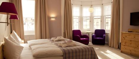 Quarto de hotel com cama, duas poltronas, cómoda de madeira, televisão e janela com cortinas.