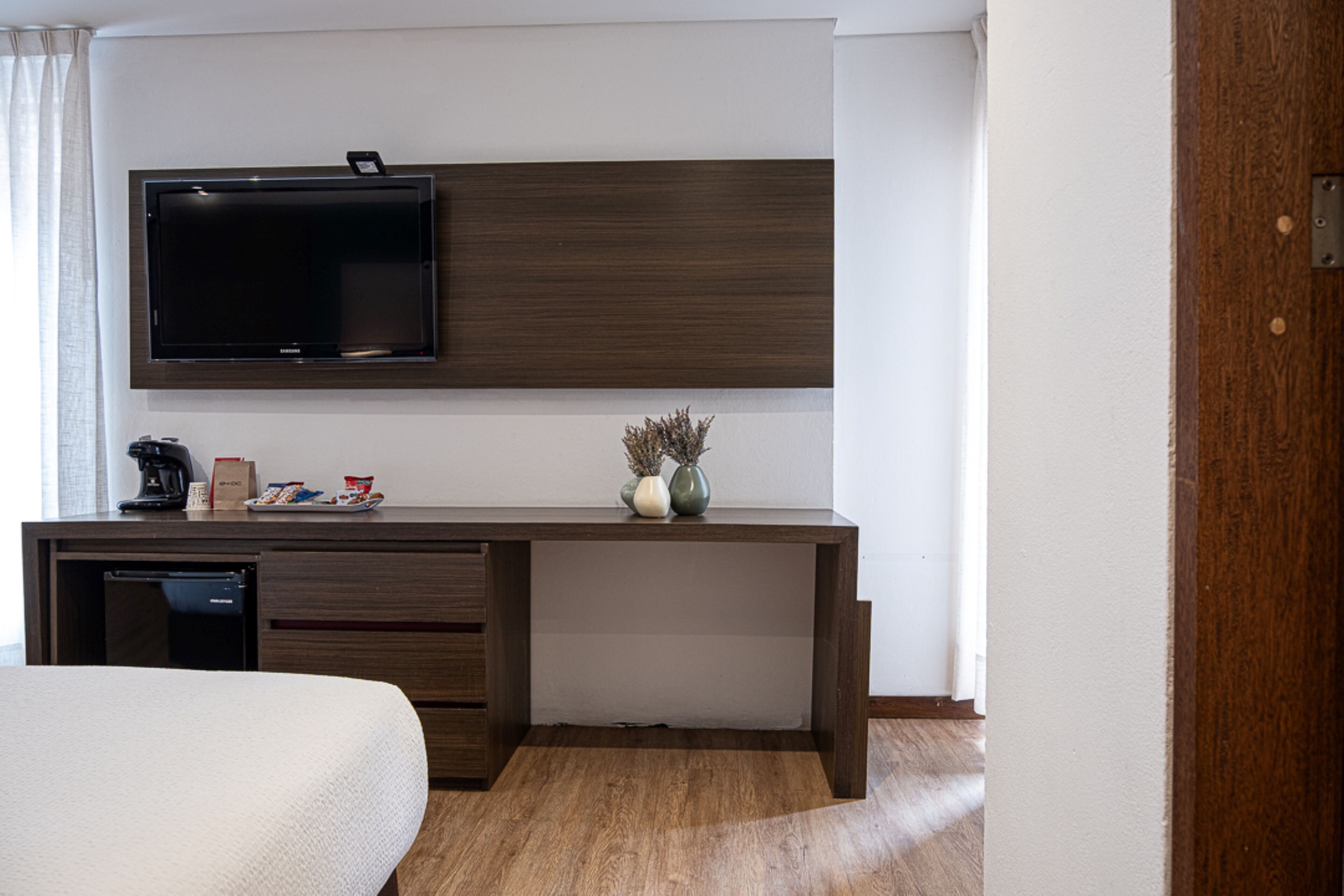 Quarto de hotel com uma cama, uma escrivaninha com televisão, um aparelho de café e um vaso com flores secas.