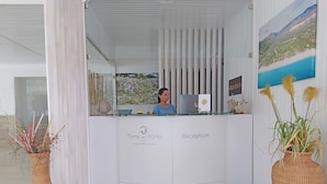 Bancada de recepção no Hotel & Residence Torre del Porto, com recepcionista, uma ampla janela e vista, e dois vasos grandes de fibra.